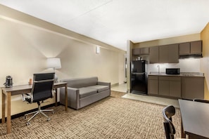 Quarto de hotel compacto com cozinha compacta, sofá, escrivaninha com luminária e uma cadeira.