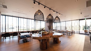 Um café amplo com grandes janelas, mesas de madeira e luminárias pendentes.