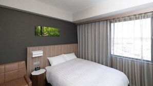 Quarto de hotel com uma cama grande, cabeceira de madeira, mesinha de cabeceira com um relógio, uma poltrona de couro marrom e uma janela com cortinas translúcidas.