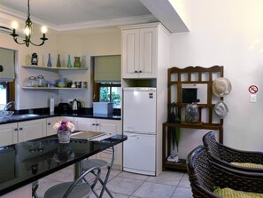 Uma cozinha com bancada preta, armários brancos e uma geladeira branca, além de um espaço de refeições com mesa de vidro e cadeiras de metal.