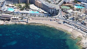 Vista aérea de um hotel à beira-mar com várias piscinas, uma praia e uma estrada que acompanha a orla.