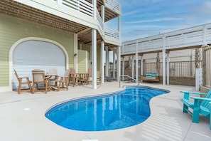 Piscina com borda curva, rodeada por cadeiras de madeira e um espaço de estar coberto.