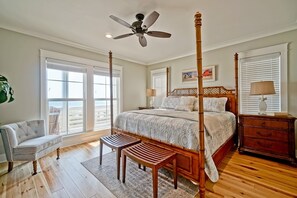 Uma cama com dossel, um ventilador de teto, um banco de madeira, uma mesa de cabeceira com abajur e vista para o oceano através de amplas janelas.