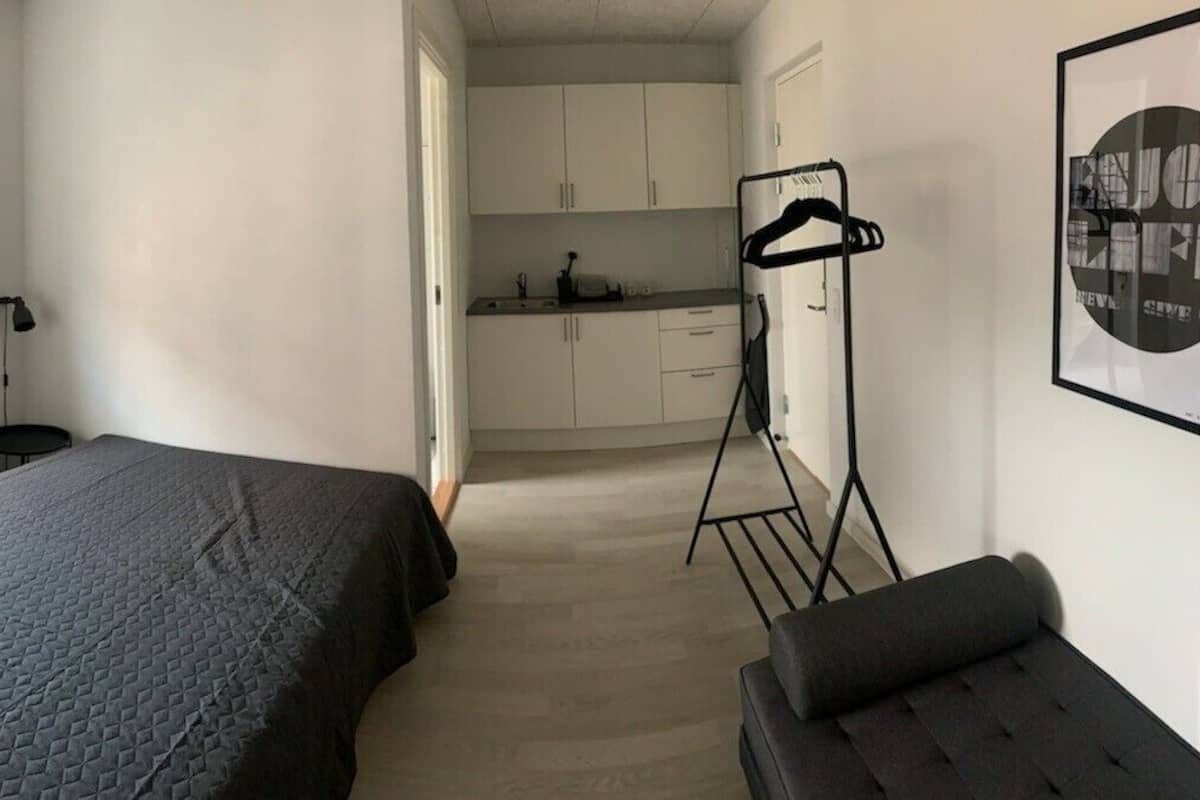 Quarto compacto com cama, mesa de cabeceira e pequena escrivaninha. Uma janela permite a entrada de luz natural e um radiador é visível na parede.