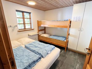Quarto em dormitório com beliches, uma janela, um guarda-roupa branco e uma pequena prateleira.