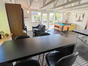 Quarto com mesa de bilhar, mesa de pebolim e mesa de sinuca. Há duas cadeiras e um sofá. O quarto tem grandes janelas e vista para o exterior.