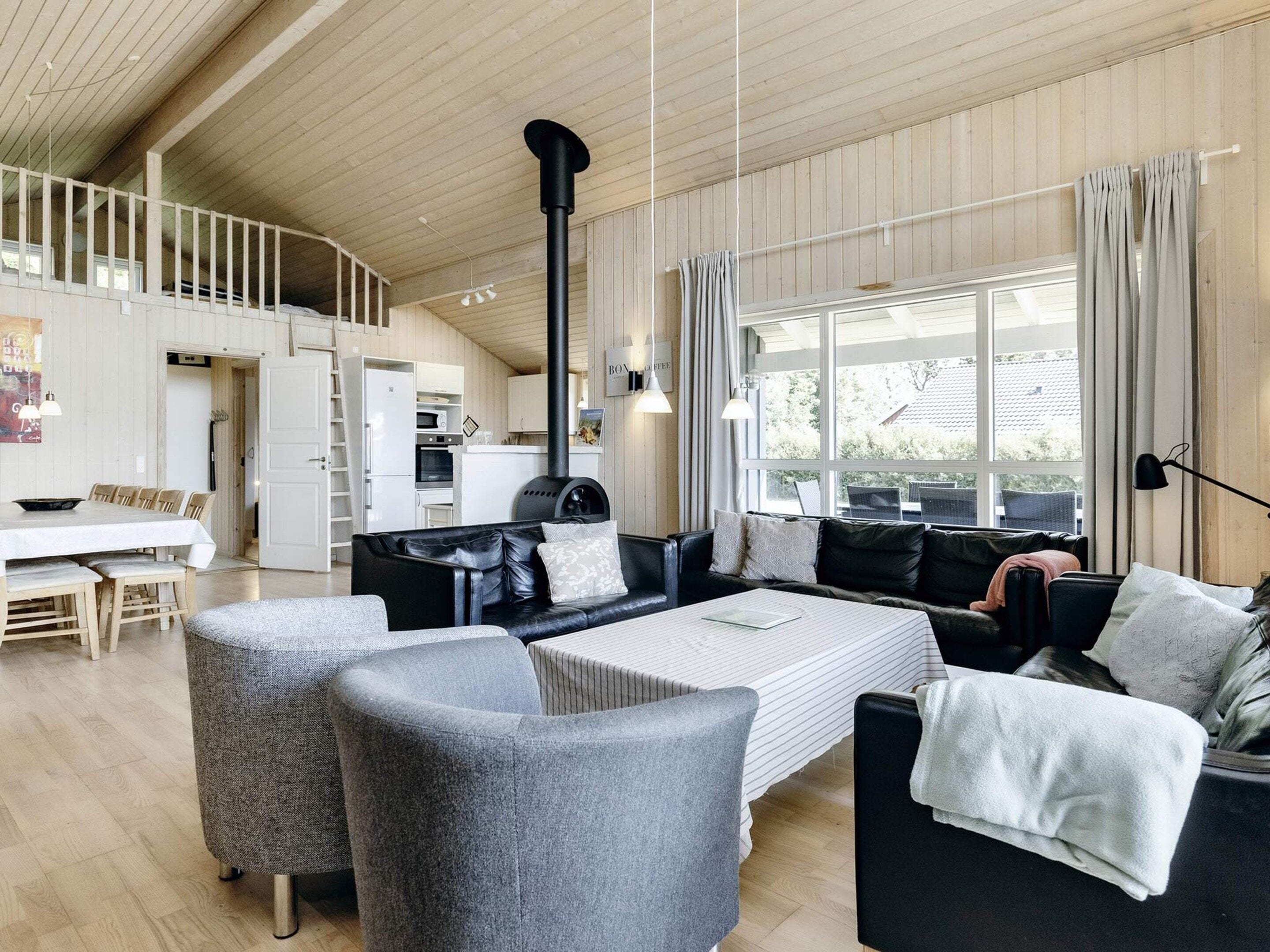 Ampla sala de estar com sofá de couro preto, poltronas cinzas e tapete listrado. Uma lareira preta está localizada no centro, e a sala possui grandes janelas com cortinas.