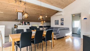 Uma cozinha moderna com vigas de madeira no teto, sala de jantar com mesa de madeira e cadeiras pretas, e um sofá cinza numa ampla sala de estar.