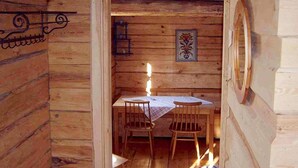 Interior de uma cabana de madeira com uma mesa de jantar e cadeiras, um pequeno armário com objetos e um detalhe decorativo na parede.