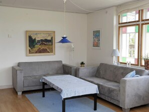 Uma sala de estar com um sofá cinzento, uma mesa pequena com toalha branca, um candeeiro pendente azul e um quadro na parede.
