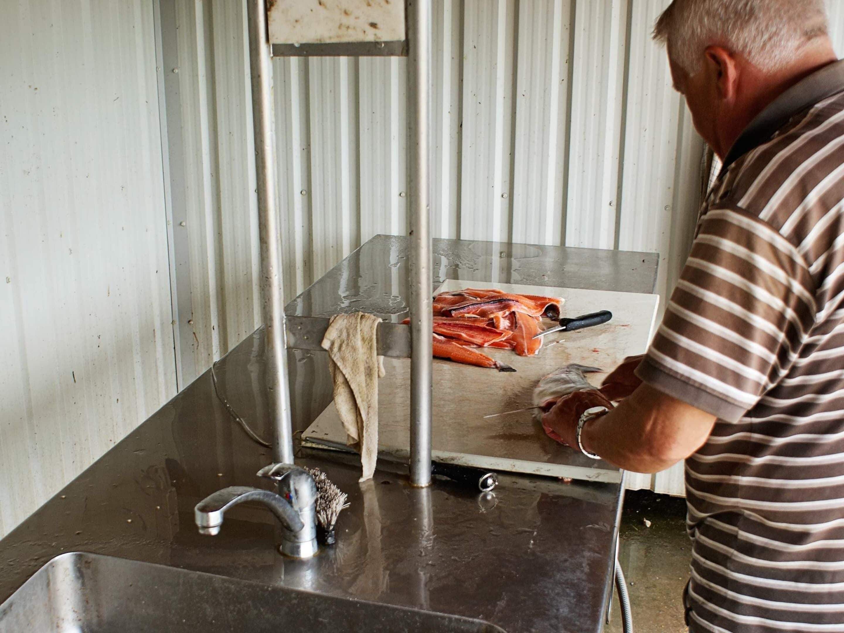 Um homem trabalhando em uma bancada de aço inoxidável, com uma faca e peixe, uma pia e uma toalha pendurada na parede.