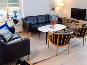 Uma sala de estar moderna com um sofá de couro preto, duas cadeiras de madeira, uma mesa de centro branca e uma televisão de tela plana.