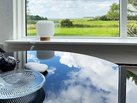 Quarto com vista para um campo verde através de uma janela, abajur branco em uma prateleira e superfície refletora com reflexo de céu azul.