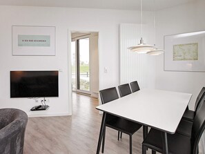 Uma área de jantar moderna com mesa branca e cadeiras pretas, televisão de tela plana na parede e luminária pendente sobre a mesa.