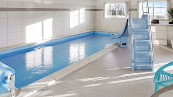 Piscina interna com escorregador, escada e sala iluminada pelo sol com janela.