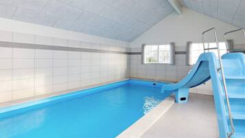 Piscina interna com escorregador azul, paredes azulejadas e janela com cortinas.