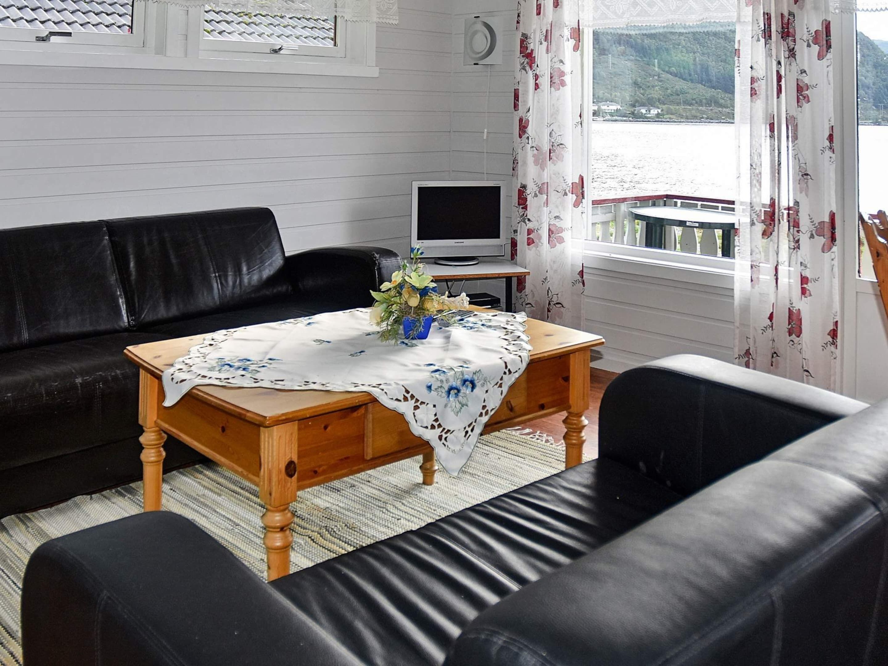 Uma sala de estar com um sofá de couro preto, uma mesa de centro de madeira com toalha florida, uma televisão em um suporte e uma janela com cortinas.