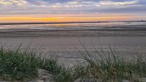 Uma praia com areia e grama ao primeiro plano, e um pôr do sol ao fundo.