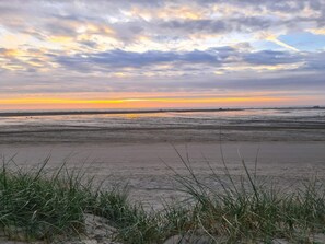 Uma praia com areia e grama ao primeiro plano, e um pôr do sol ao fundo.