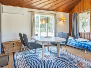 Um quarto com uma mesa redonda, duas cadeiras, uma cama com colcha azul, um abajur e uma janela com cortinas.