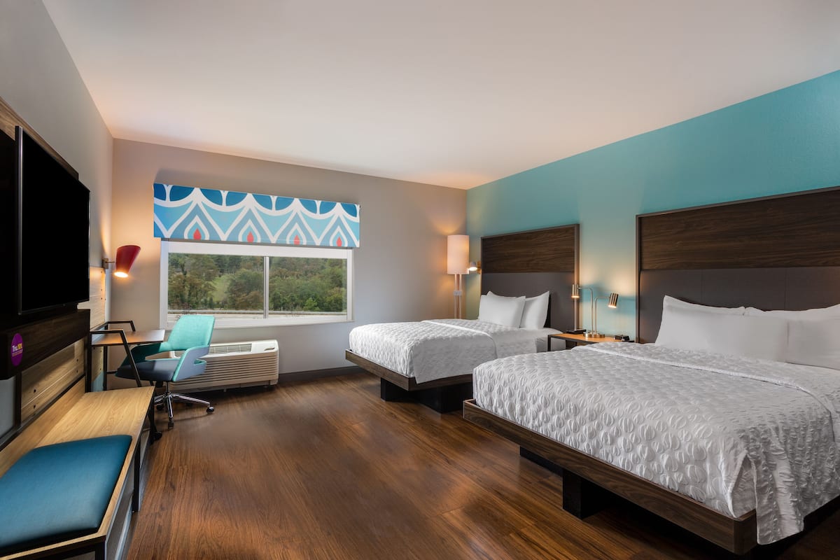 Quarto de hotel com duas camas, uma escrivaninha, uma cadeira, uma televisão e uma janela com vista para o verde.