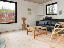 Uma sala de estar com uma mesa de centro de madeira, um sofá de couro preto, uma poltrona de vime e vista para um prédio vermelho através de uma janela grande.