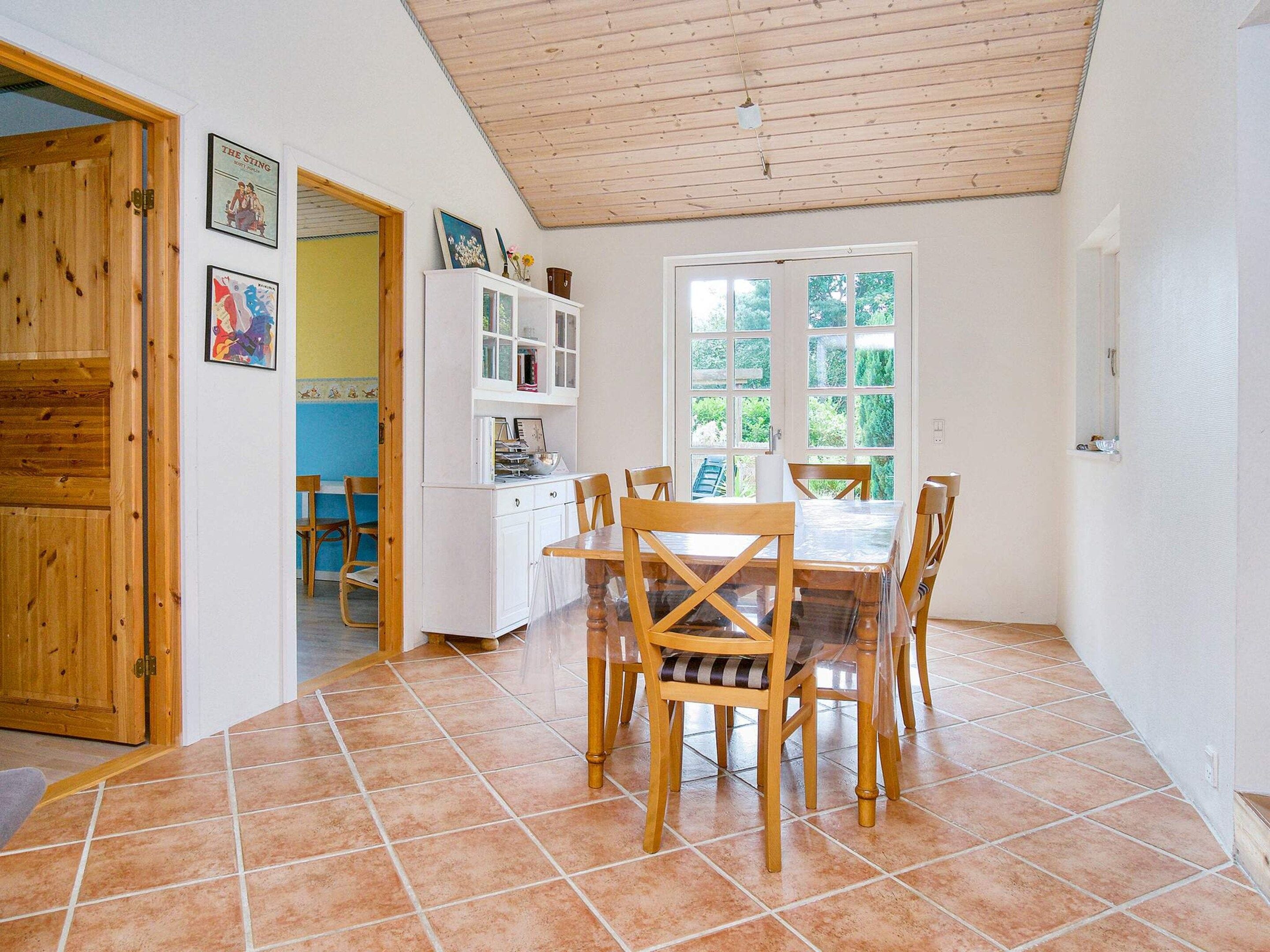 Uma área de refeições com mesa e cadeiras de madeira, um armário branco com portas de vidro e vista para o verde através de uma grande janela.