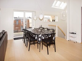 Uma área de refeições com uma mesa redonda, cadeiras pretas e luminárias pendentes. Há uma escada com corrimão e uma parede branca com um objeto decorativo.
