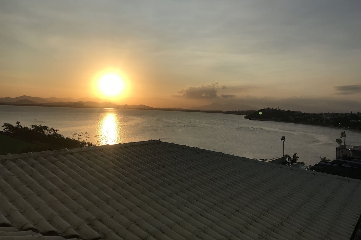 Um pôr do sol sobre um rio calmo, com um telhado de telhas ao primeiro plano.