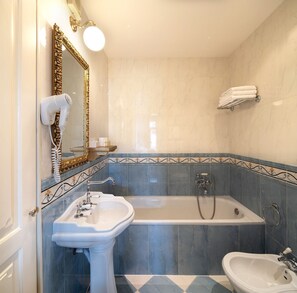 Um banheiro com banheira, pia e bidê. Há um espelho com moldura decorativa, um porta-toalhas e um secador de cabelo fixado na parede.
