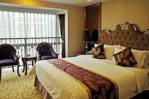 Quarto de hotel com uma cama grande, duas poltronas, uma mesa pequena e vista para o exterior através de cortinas leves.