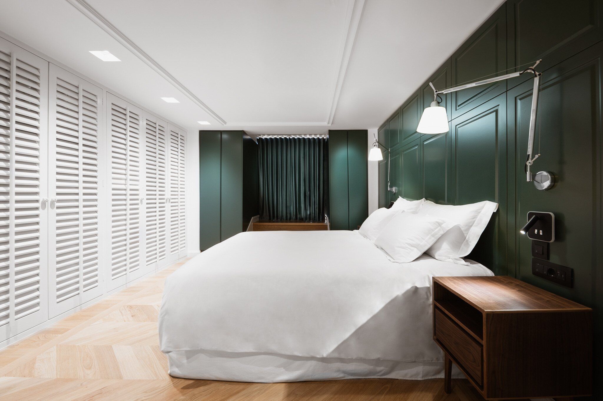 Quarto moderno com cama grande, mesa de cabeceira de madeira e luminária fixada na parede.