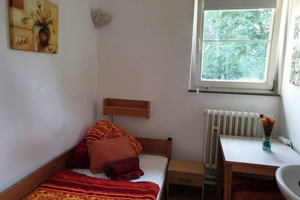 Quarto pequeno e simples, com uma cama, uma escrivaninha, um radiador e uma janela com vista para árvores.