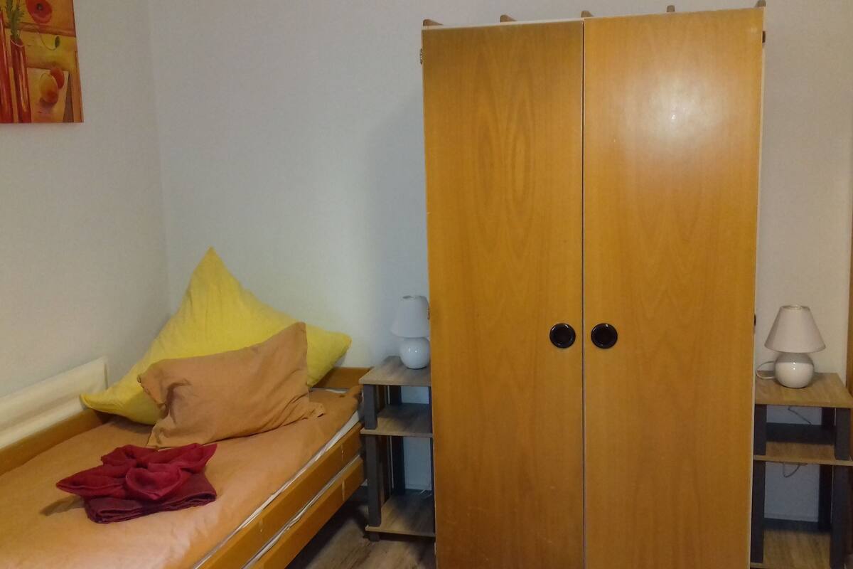 Quarto com uma cama, guarda-roupa de madeira, mesa de cabeceira com abajur e um quadro na parede.