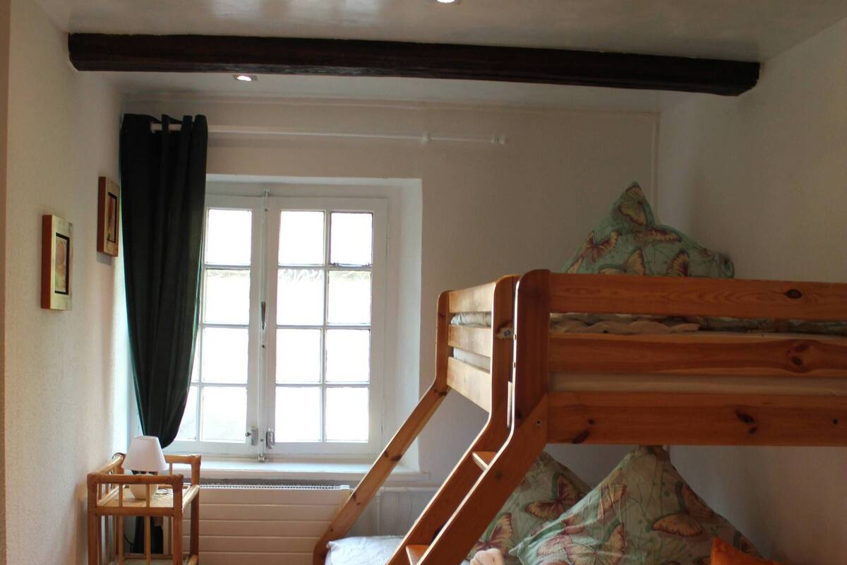 Uma cama beliche com estrutura de madeira, uma mesinha com cadeira e uma janela com cortinas.