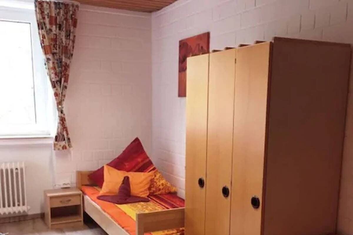 Quarto pequeno, com uma cama de solteiro, guarda-roupa de madeira e janela com cortinas.