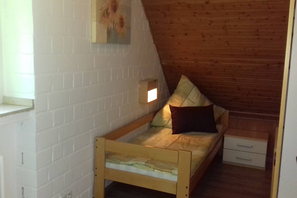 Um quarto pequeno e acolhedor com teto inclinado, uma cama de madeira com almofadas, uma mesa de cabeceira pequena e uma janela com vista para o exterior.