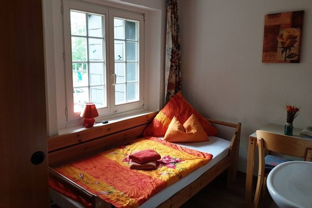 Uma cama de solteiro com um colcha colorida, uma janela com cortinas, uma pequena mesa com um vaso e uma cadeira.