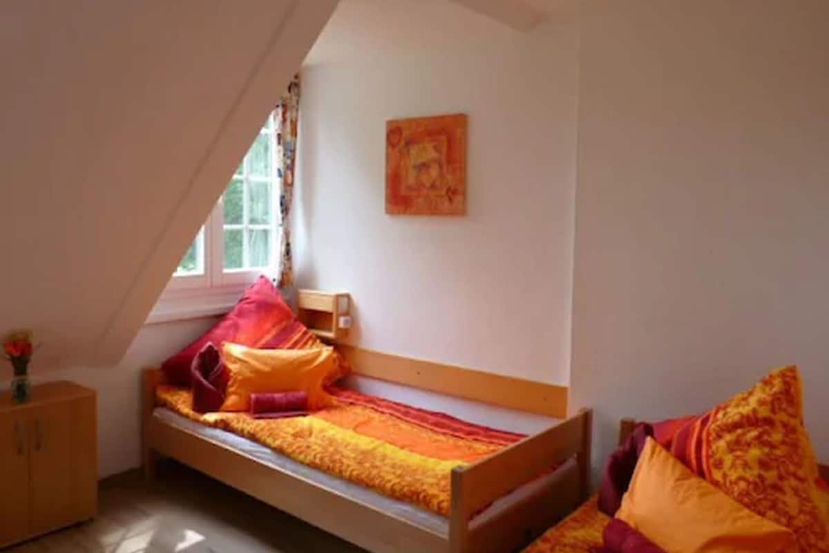 Um quarto pequeno e acolhedor com teto inclinado, uma cama de solteiro com roupa de cama laranja, uma mesinha de cabeceira de madeira e uma janela com cortinas.