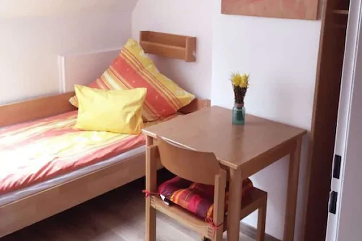 Um quarto pequeno e aconchegante com uma cama de solteiro, uma escrivaninha de madeira e uma cadeira. Há uma pequena mesa com um vaso de flores.