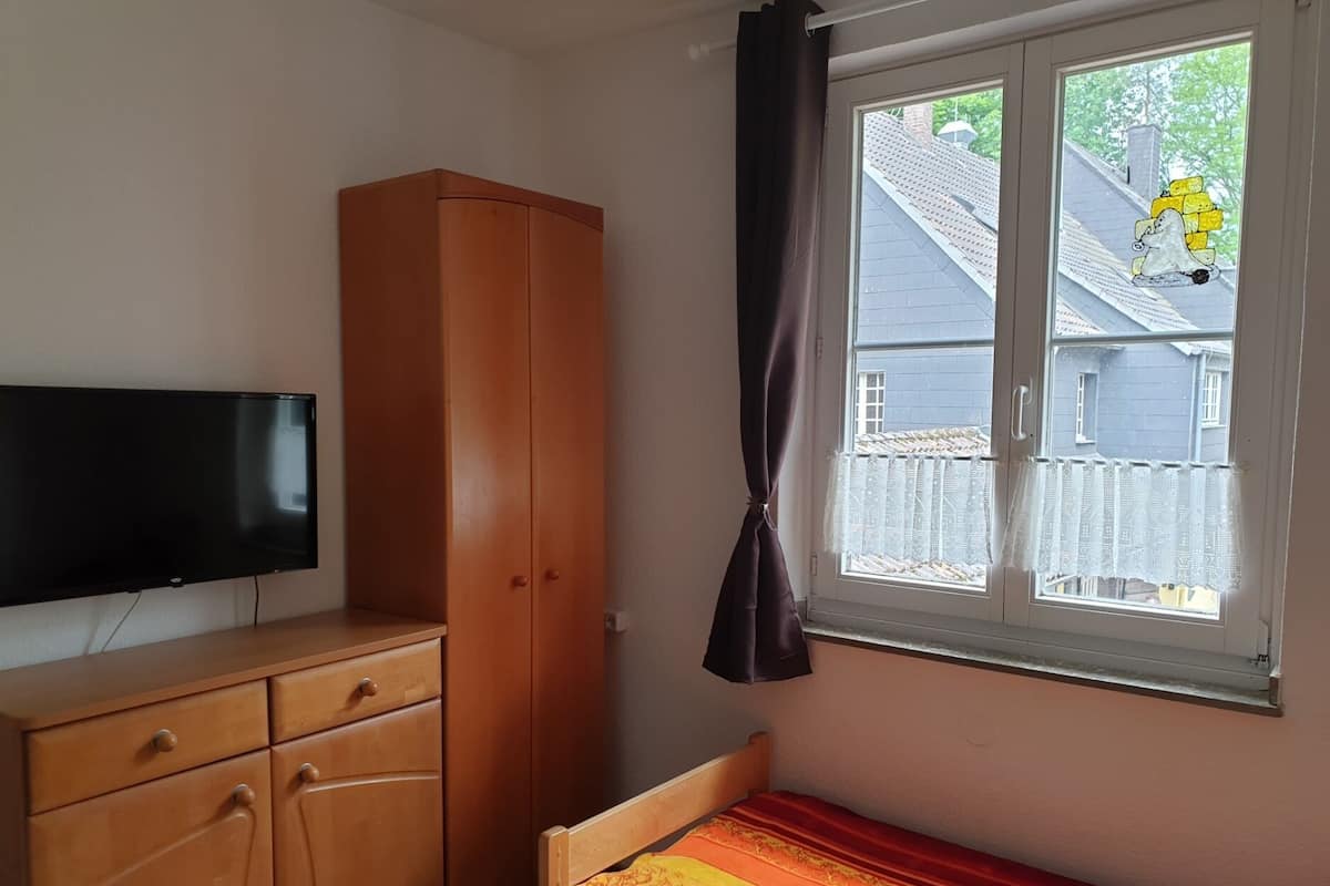 Quarto com cama, guarda-roupa de madeira, televisão na parede e janela com vista para uma casa.