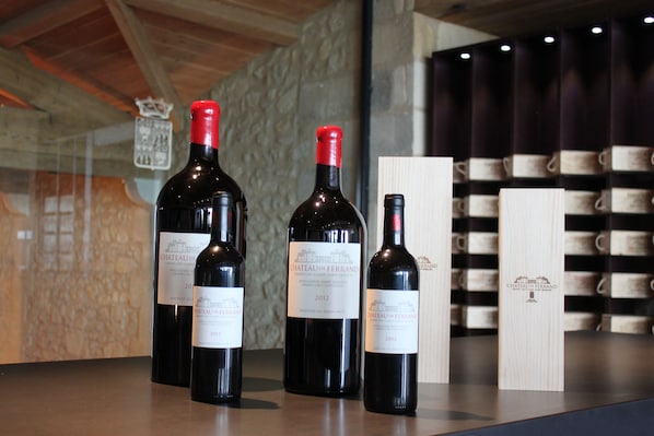 Três garrafas de vinho Chateau de Ferrand sobre um balcão, com uma parede de pedra e teto de madeira ao fundo.