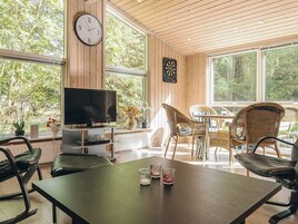 Quarto com televisão, cadeiras de vime, mesa de jantar e janelas amplas.