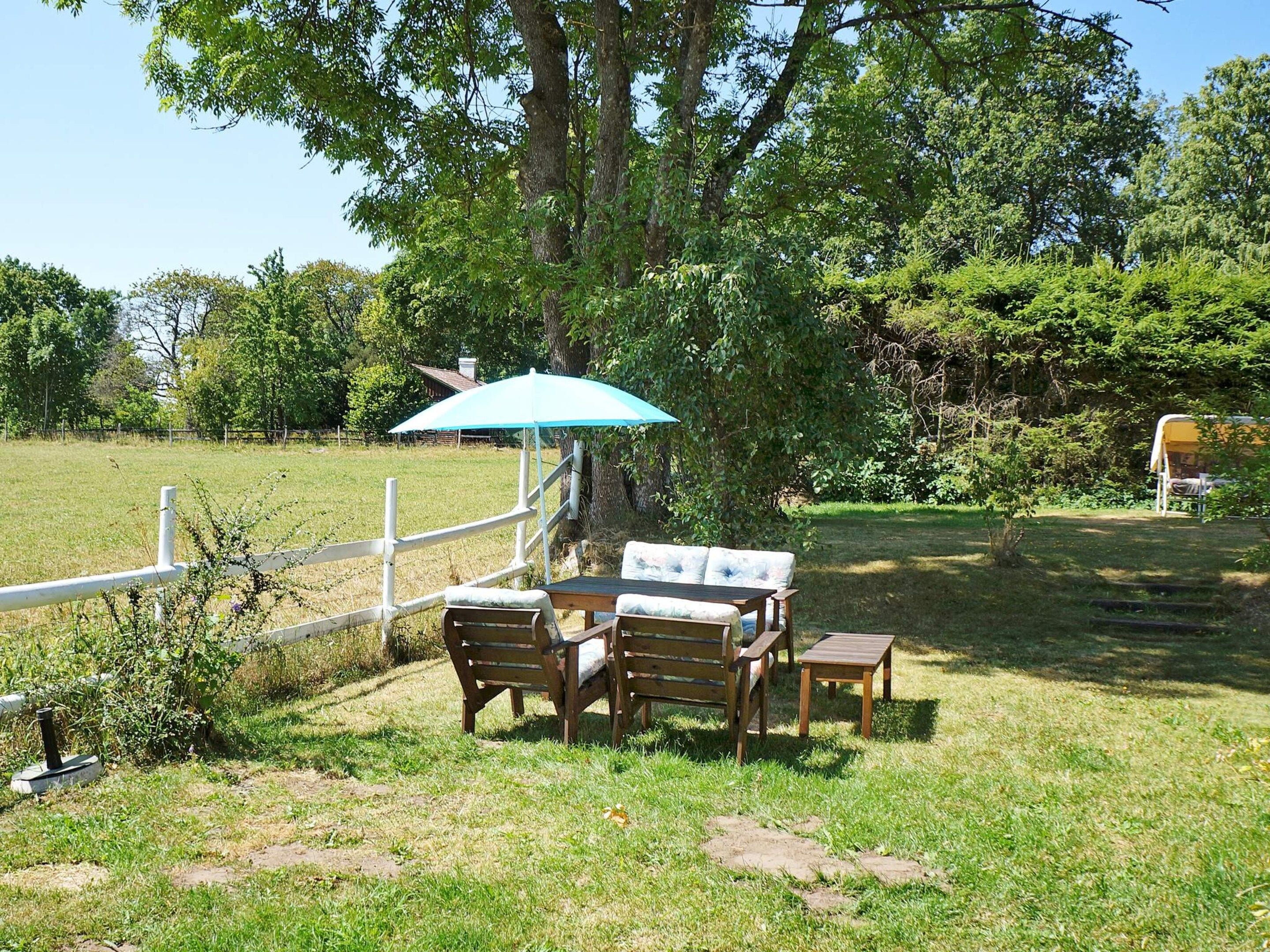 Um espaço para piquenique com mesa, cadeiras e um guarda-sol azul, cercado por gramado e árvores.