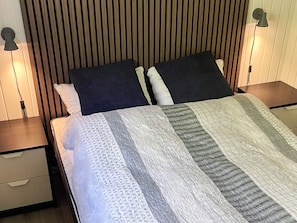Cama bem arrumada com manta estampada, dois travesseiros, cabeceira de madeira e abajures laterais.