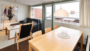 Uma sala de estar moderna com um sofá, uma mesa de jantar e cadeiras. Há uma janela grande com cortinas, um quadro na parede e um abajur.