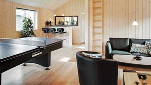 Quarto com mesa de ping pong, sofá, mesa de centro, escada e área de cozinha com banquetas.