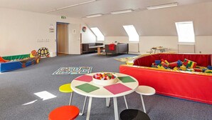 Uma sala de jogos ampla com teto alto, claraboias e diversos equipamentos de recreação, incluindo uma área de atividades em piso macio e uma mesa com tapetes coloridos.