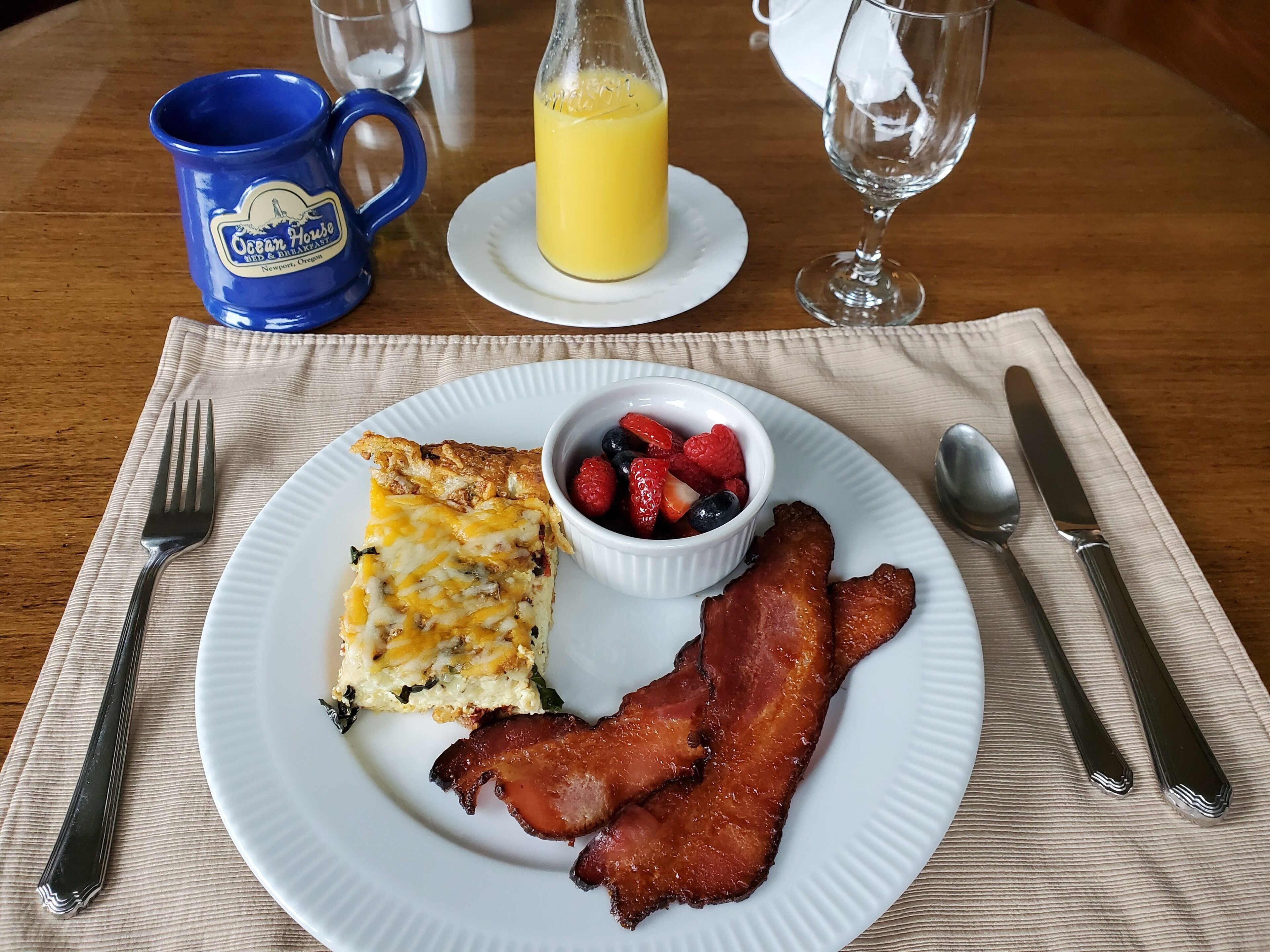 Prato de café da manhã com bacon, uma fatia de quiche e uma tigela de frutas vermelhas.