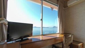 Quarto com escrivaninha, cadeira, televisão e uma grande janela com vista para o mar.